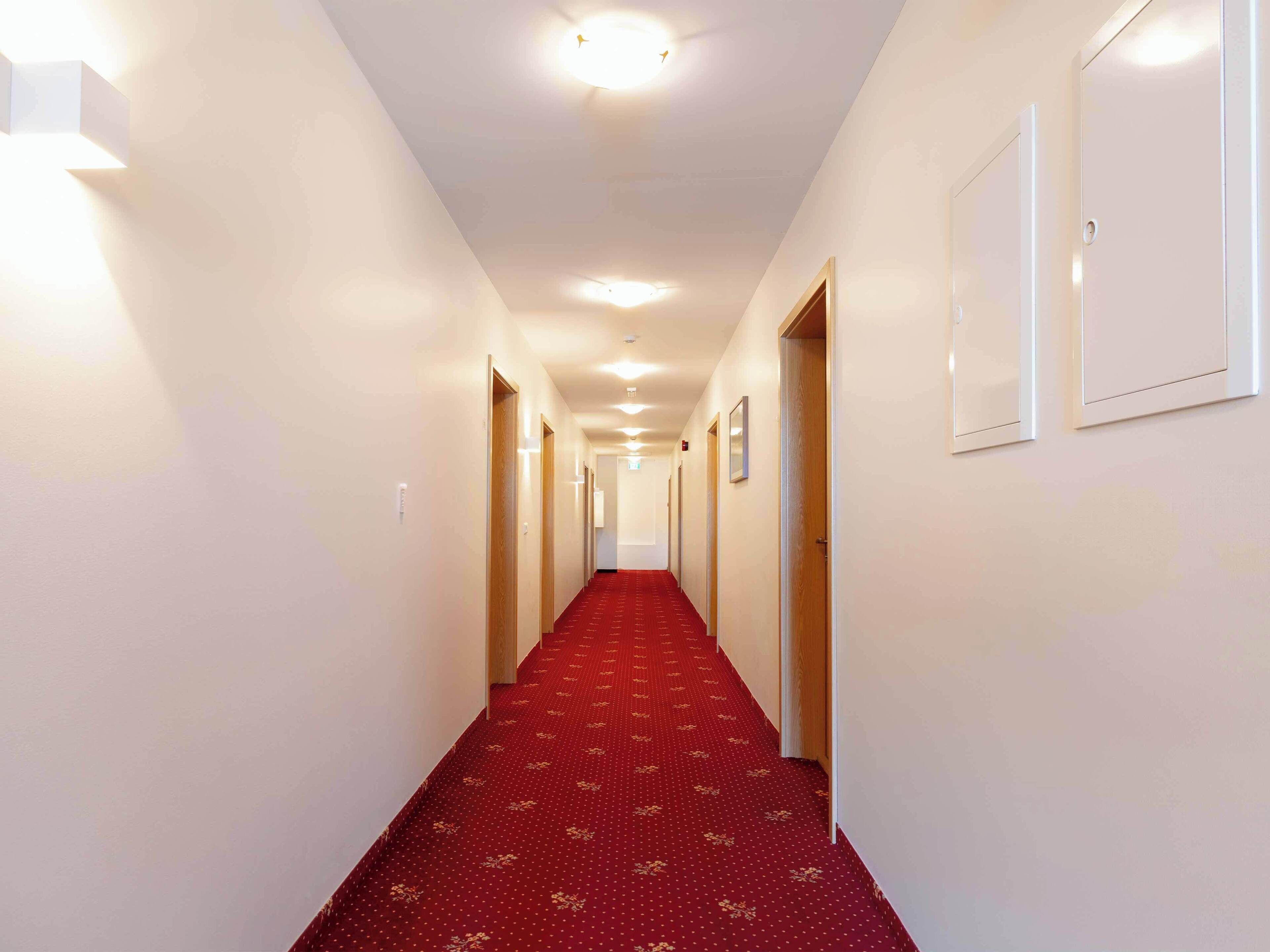 Corredor de hotel com tapete vermelho, paredes brancas e portas de acesso aos quartos.