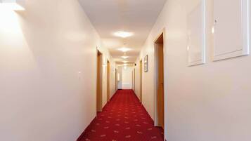 Corredor de hotel com tapete vermelho, paredes brancas e portas de acesso aos quartos.