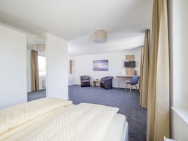 Quarto de hotel com uma cama, duas cadeiras, uma escrivaninha, televisão e um quadro na parede.