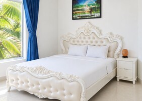 Uma cama branca com cabeceira estofada, uma mesinha de cabeceira com um vaso e um quadro na parede.
