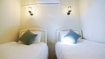 Duas camas de solteiro com cabeceiras, roupa de cama branca e duas luminárias fixadas na parede.