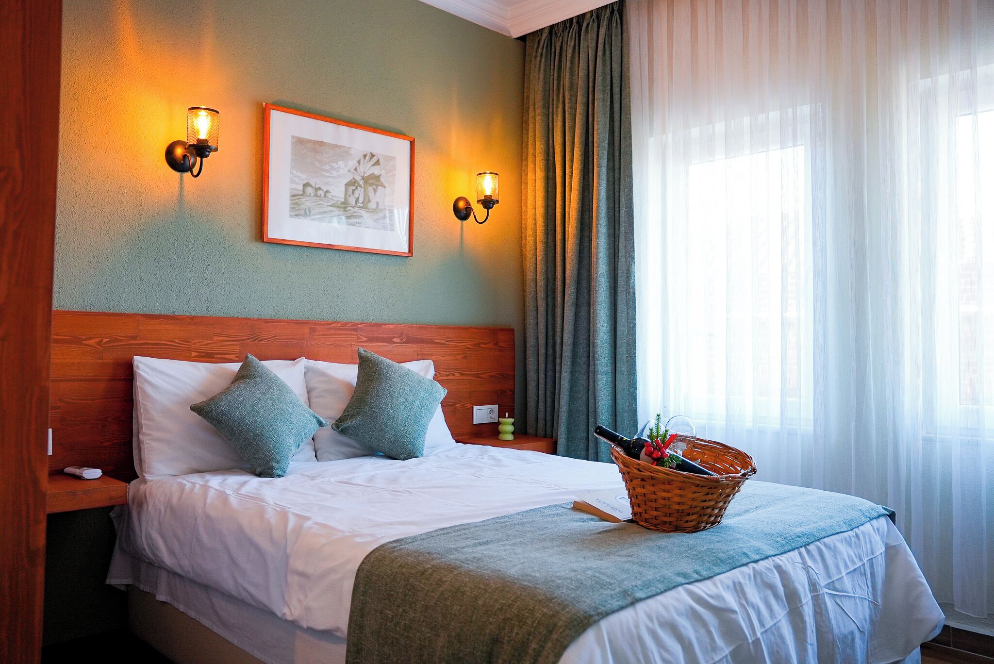 Quarto de hotel com uma cama, duas arandelas de parede, um quadro emoldurado, um cesto com objetos e uma janela com cortinas.