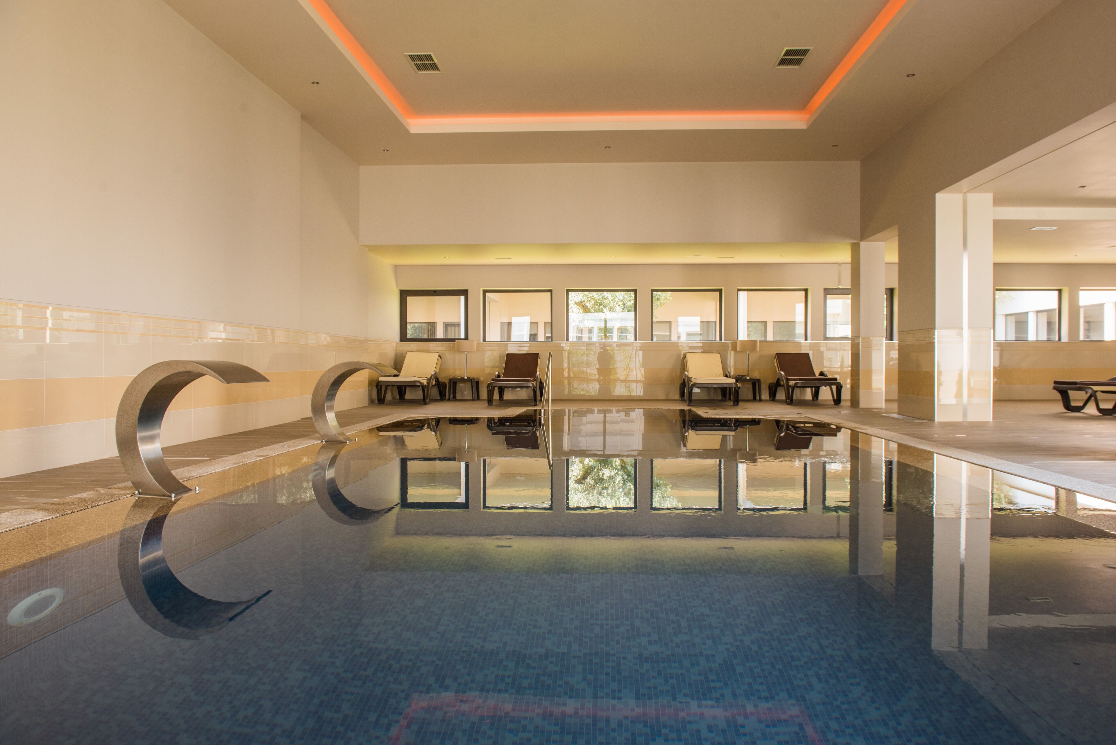 Uma piscina interior moderna com água cristalina, cadeiras de descanso e iluminação laranja no teto.