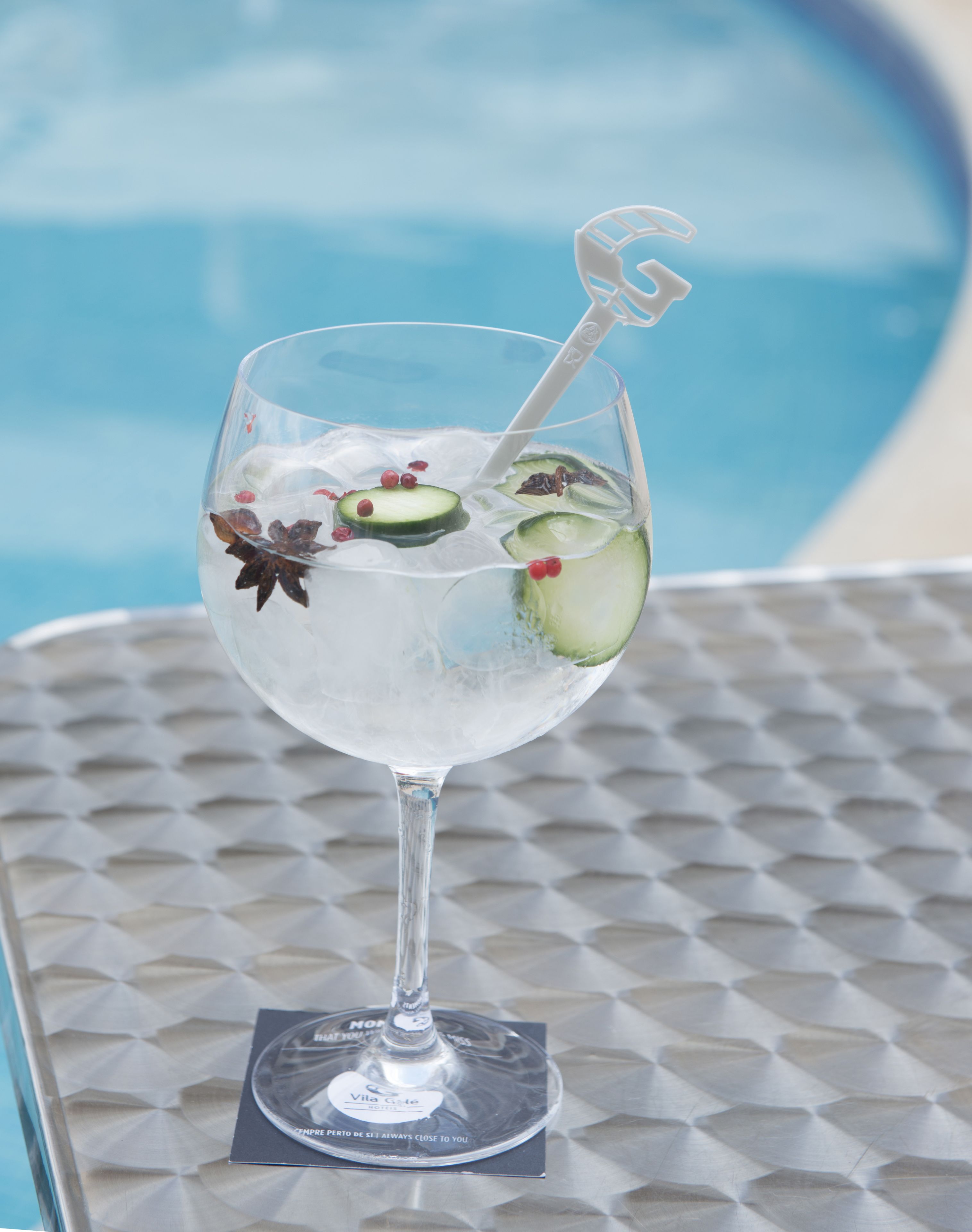 Um copo de vidro transparente com gelo, fatias de pepino e anis estrelado, sobre uma mesa com textura perto de uma piscina.