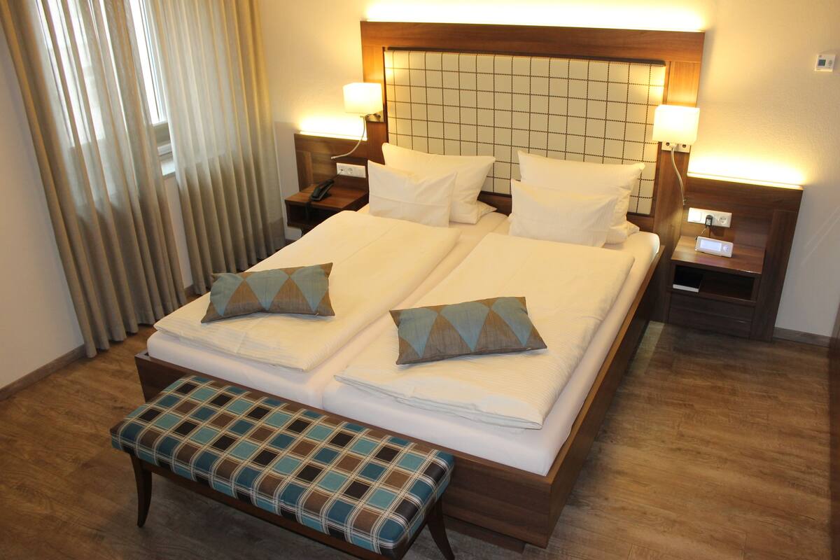 Uma cama de casal com duas almofadas, um banco e duas mesas de cabeceira com abajures.