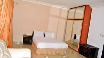 Quarto de hotel com cama, mesa de cabeceira, espelho, mesa de centro de vidro e sofá branco.