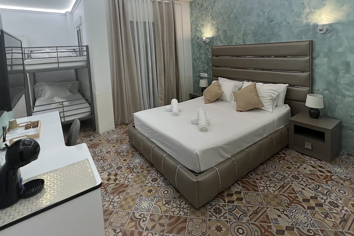 Quarto de hotel com uma cama de solteiro, escrivaninha com cadeira, mesa de cabeceira com abajur e ladrilhos no chão com padrão.