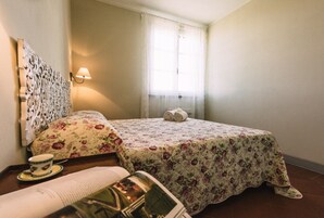 Quarto com colcha florida, mesa de cabeceira com xícara e pires, e luminária na parede.