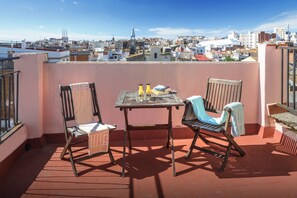 Uma varanda com duas cadeiras de madeira, uma mesa pequena e vista para o horizonte urbano.