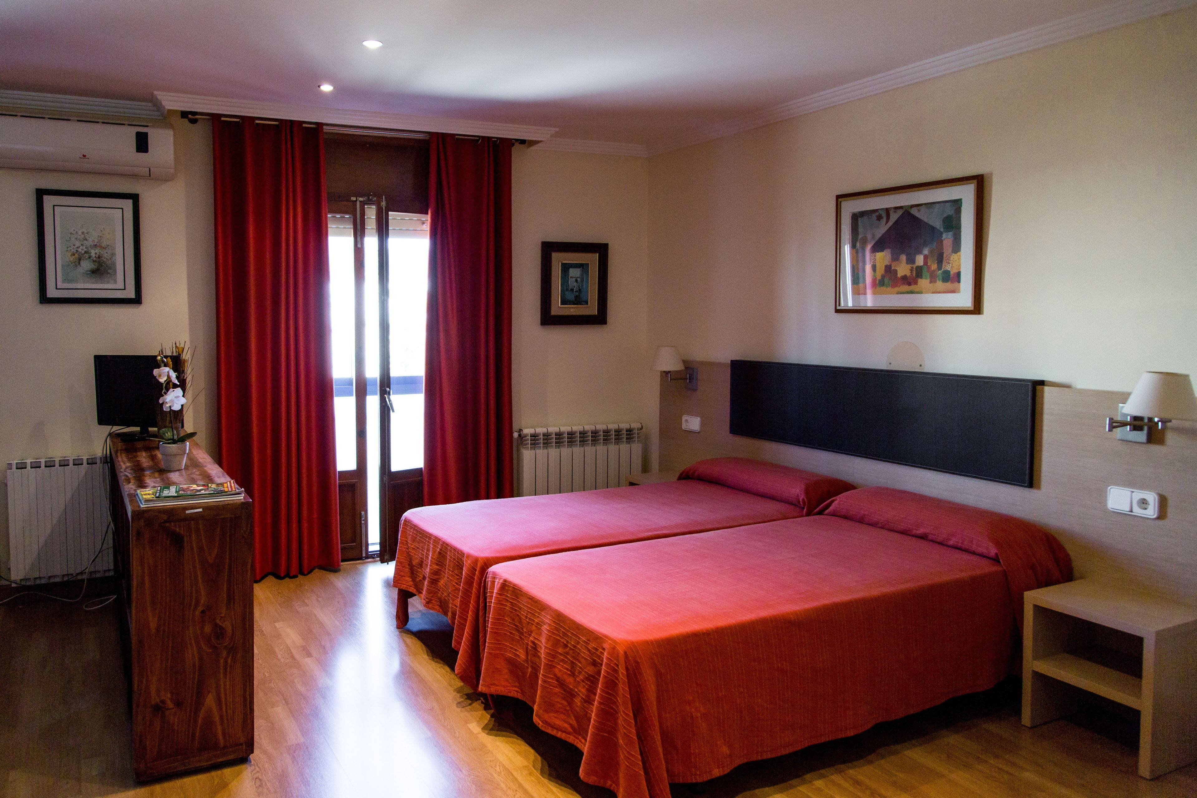 Quarto de hotel com duas camas, uma escrivaninha de madeira, um radiador e uma janela com cortinas vermelhas.