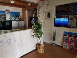 Uma área de recepção com balcão de madeira, uma planta pendente, uma TV fixada na parede e um cartaz divulgando vinho da Sardenha.