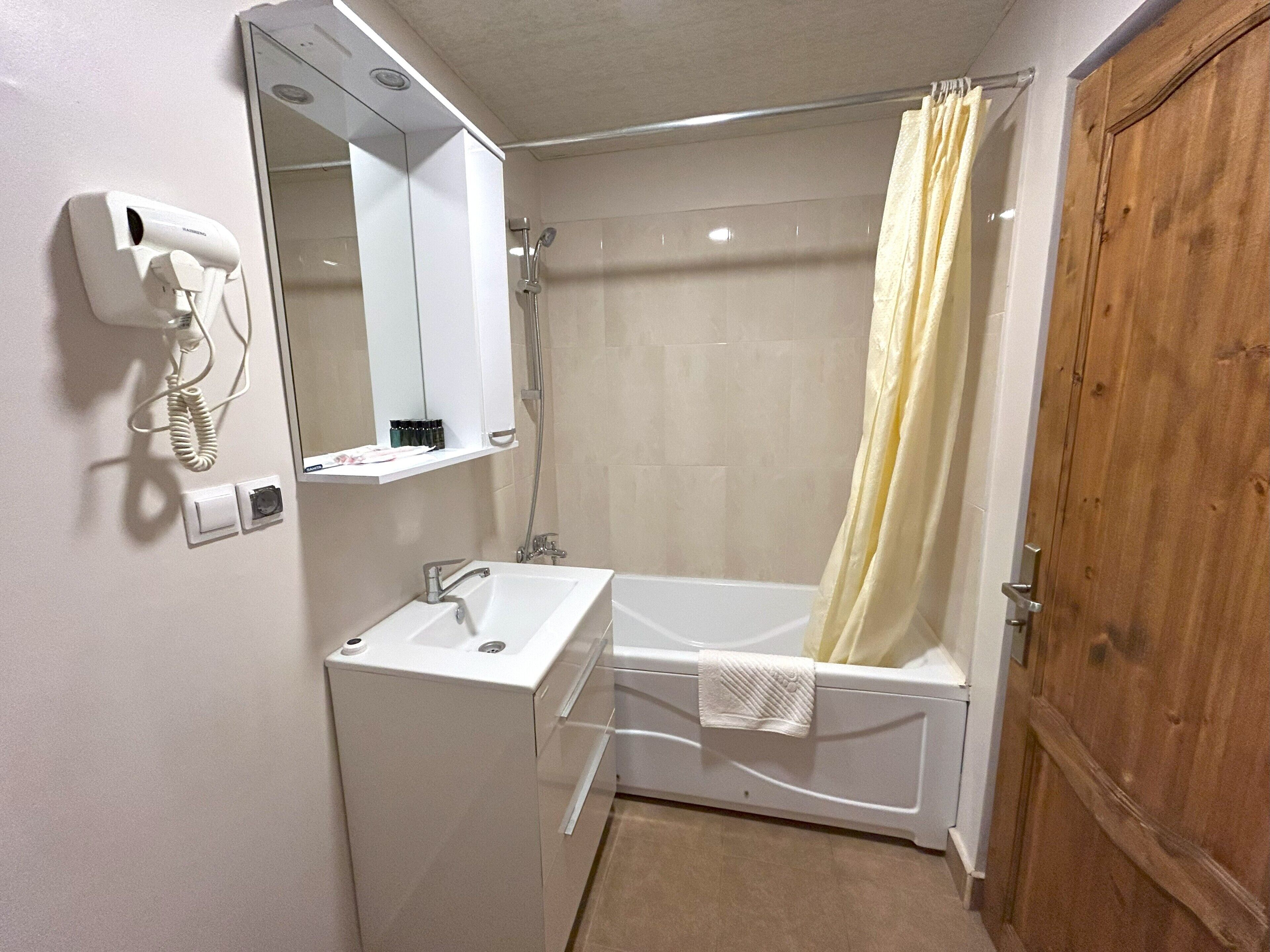 Uma casa de banho com uma pia branca, um espelho, um secador de cabelo, chuveiro com cortina amarela e uma porta de madeira.