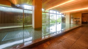 Uma piscina interior ampla com paredes de vidro transparente, chão ladrilhado e teto com luminárias lineares.