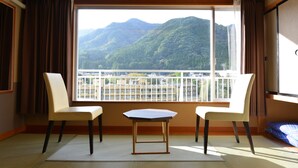 Quarto com duas cadeiras, uma mesa pequena e uma janela grande com vista para montanhas e uma ponte.