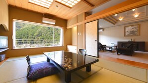 Um quarto tradicional japonês com tatame, uma mesa baixa com almofada, uma porta deslizante, um sofá e vista para uma área arborizada através de uma grande janela.