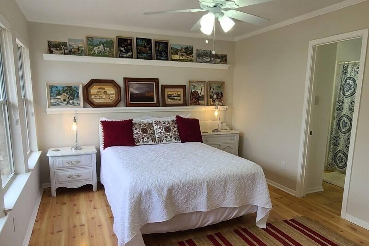 Um quarto com uma cama grande, piso de madeira, um ventilador de teto e obras de arte na parede.