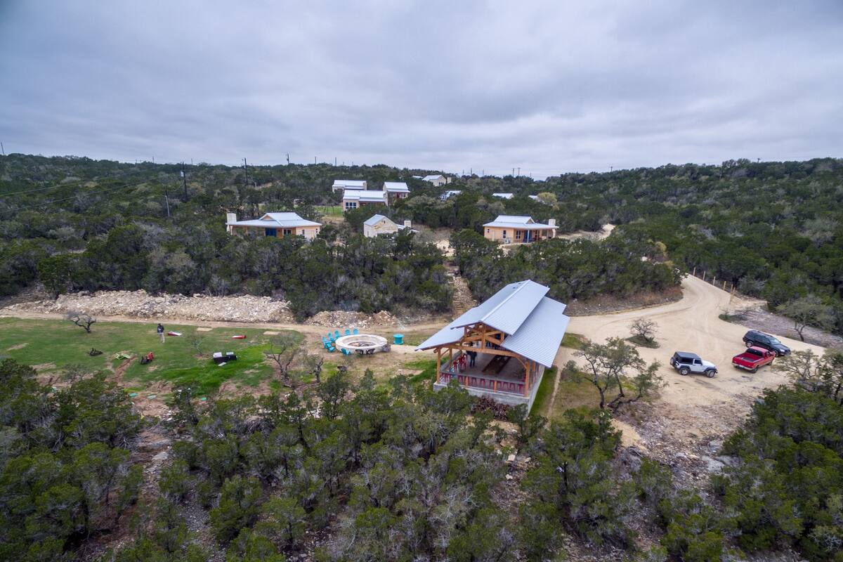 Uma pousada rústica e isolada, com várias cabanas cercadas por floresta densa, um caminho de terra e uma ampla área aberta com assentos externos.