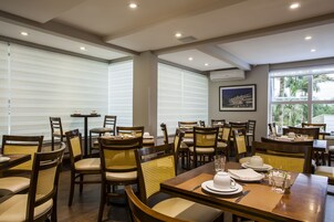 Um restaurante com mesas e cadeiras de madeira, dispostas num espaço amplo com grandes janelas e um aparelho de ar condicionado na parede.