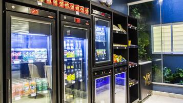 Uma fileira de refrigeradores de bebidas com diversas opções, uma planta e uma máquina de vendas automática.