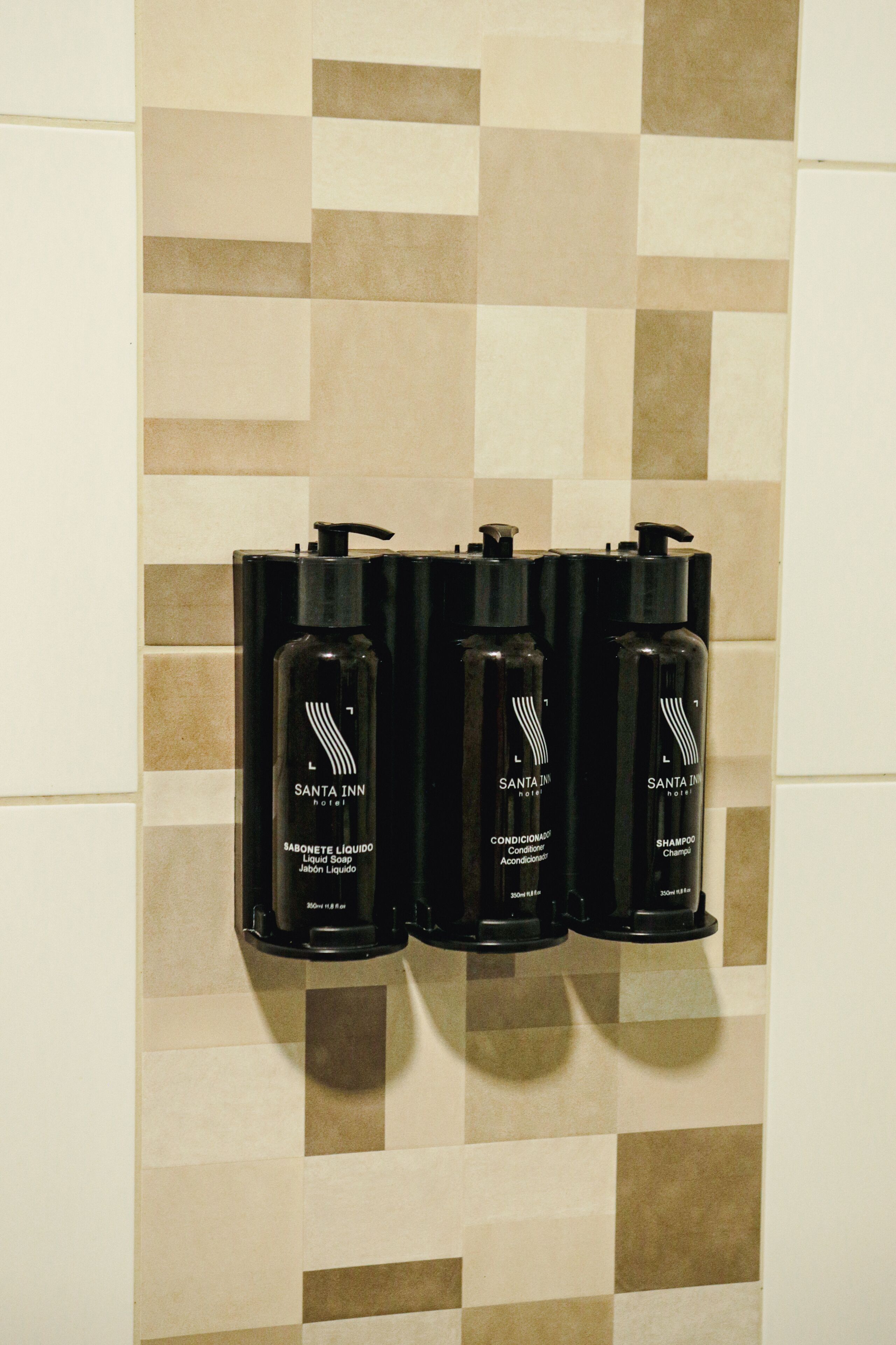 Tr ês frascos pretos de produtos de higiene pessoal do hotel Santa Inn fixados numa parede azulejada.