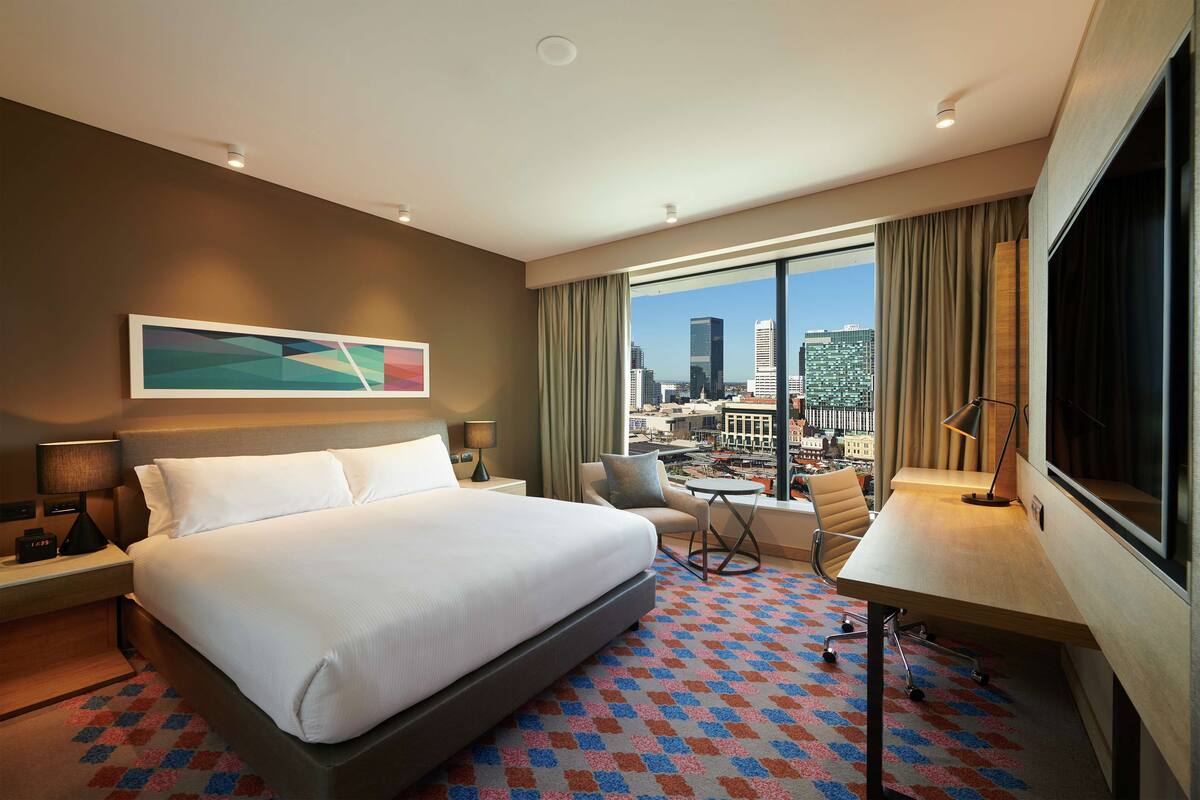 Quarto de hotel com uma cama grande, uma escrivaninha, uma cadeira e vista para a paisagem urbana.