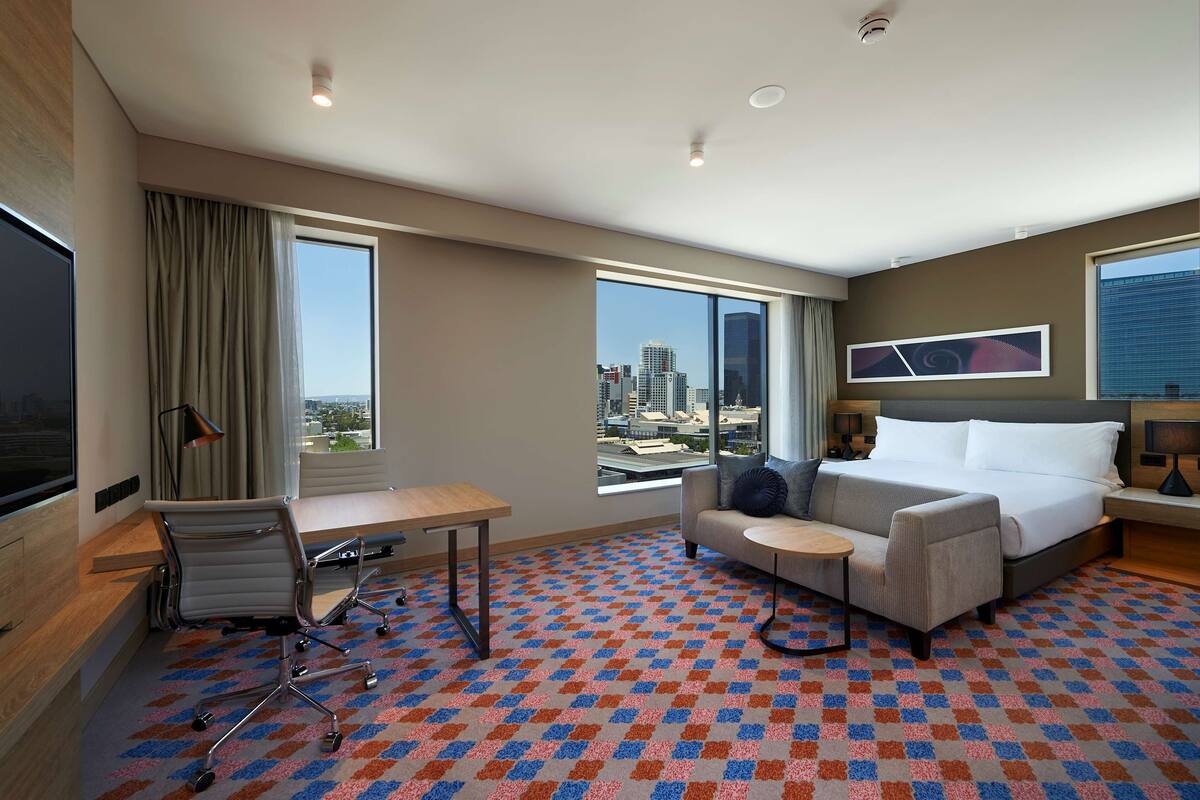 Quarto de hotel com cama grande, escrivaninha, cadeira, mesinha e televisão. Grandes janelas oferecem vista para a cidade.