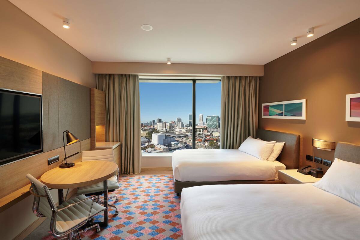 Quarto de hotel com uma grande janela, duas camas, uma escrivaninha e uma cadeira.
