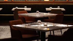 Um restaurante com iluminação suave, mesas e cadeiras, uma parede escura com prateleiras exibindo diversos objetos e um tapete estampado.