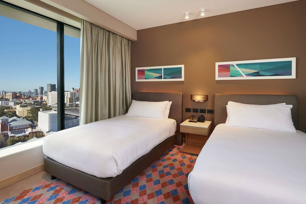 Quarto de hotel com duas camas, vista para a cidade e obras de arte nas paredes.