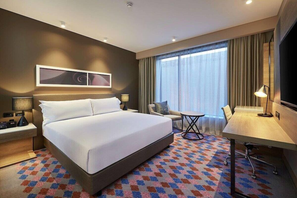 Quarto de hotel com uma cama grande, uma escrivaninha, uma cadeira, uma mesinha, uma televisão e uma janela com cortinas.