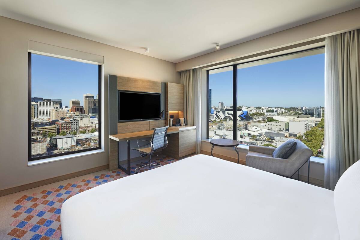Quarto de hotel com uma cama grande, uma escrivaninha com cadeira, televisão e vista para o horizonte urbano através de amplas janelas.