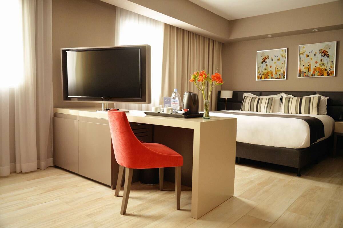 Quarto de hotel moderno com uma televisão de tela plana grande, uma escrivaninha com cadeira, uma cama com roupa de cama listrada e duas obras de arte na parede.
