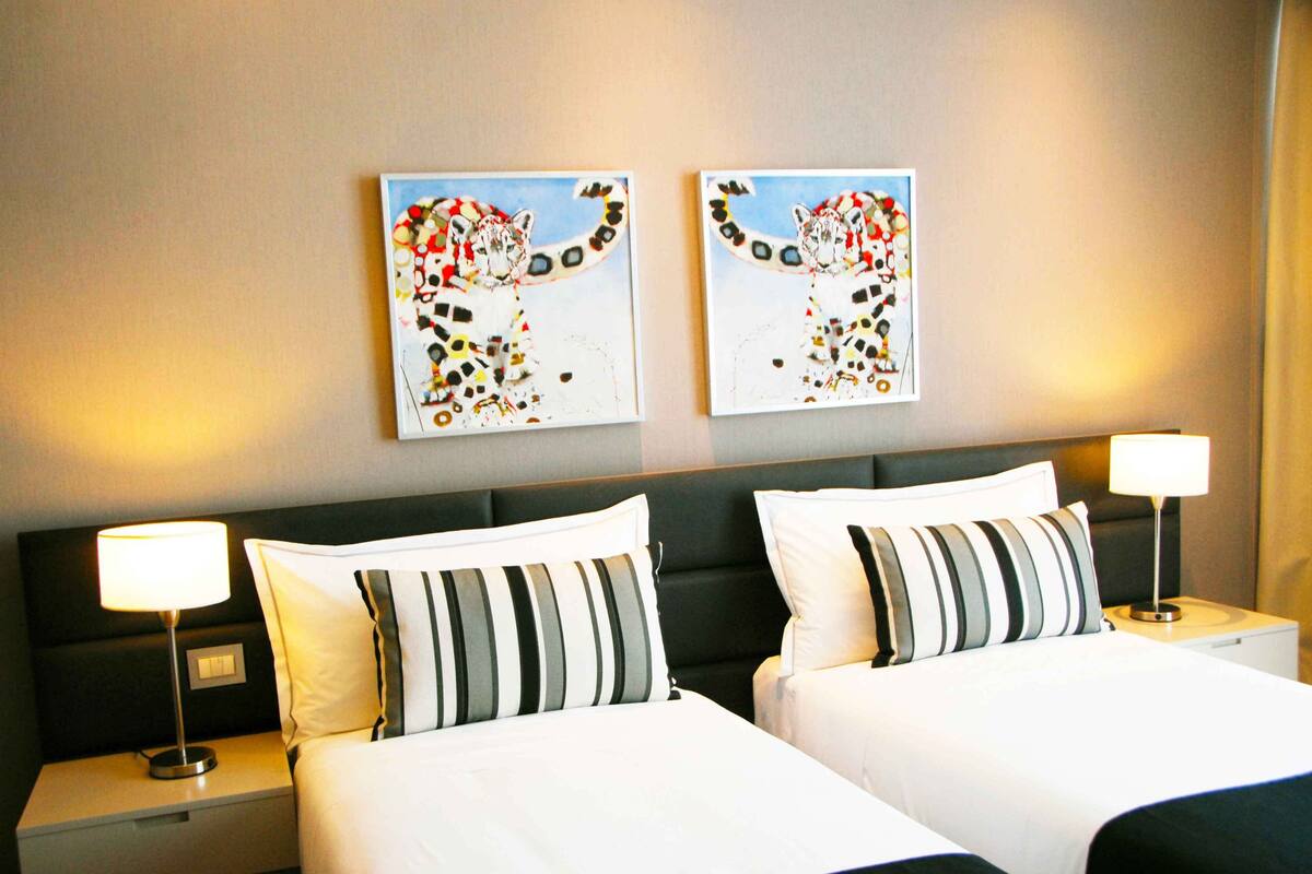 Quarto de hotel com duas camas, cada uma com almofada listrada e mesa de cabeceira com abajur. Há duas obras de arte fixadas na parede, com designs abstratos.