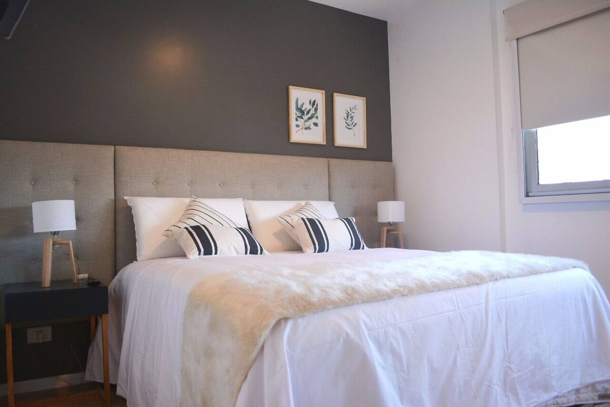 Cama bem arrumada com roupa de cama branca e almofadas listradas, com mesas de cabeceira com abajures, em um quarto com cabeceira cinza e duas gravuras botânicas emolduradas na parede.