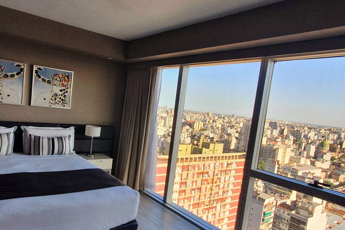 Quarto de hotel moderno com uma cama grande, duas obras de arte na parede, uma mesa de cabeceira e vista para a paisagem urbana através de grandes janelas.