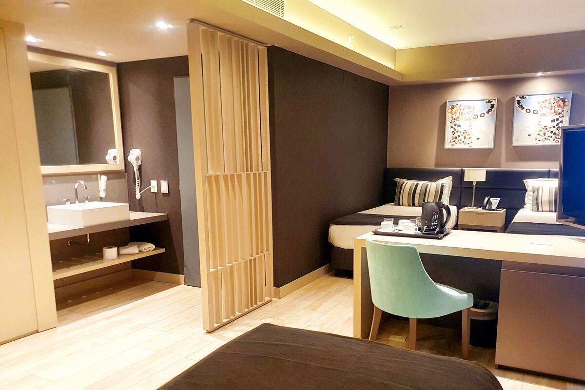 Quarto de hotel moderno com cama, escrivaninha com cadeira, banheiro com pia e espelho, e obras de arte nas paredes.