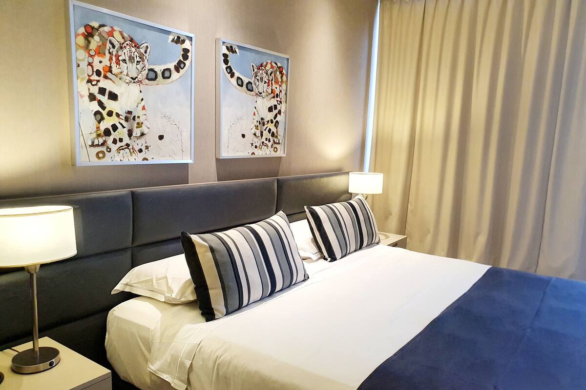 Quarto de hotel com uma cama, duas almofadas, mesa de cabeceira com abajur e duas obras de arte abstratas na parede.