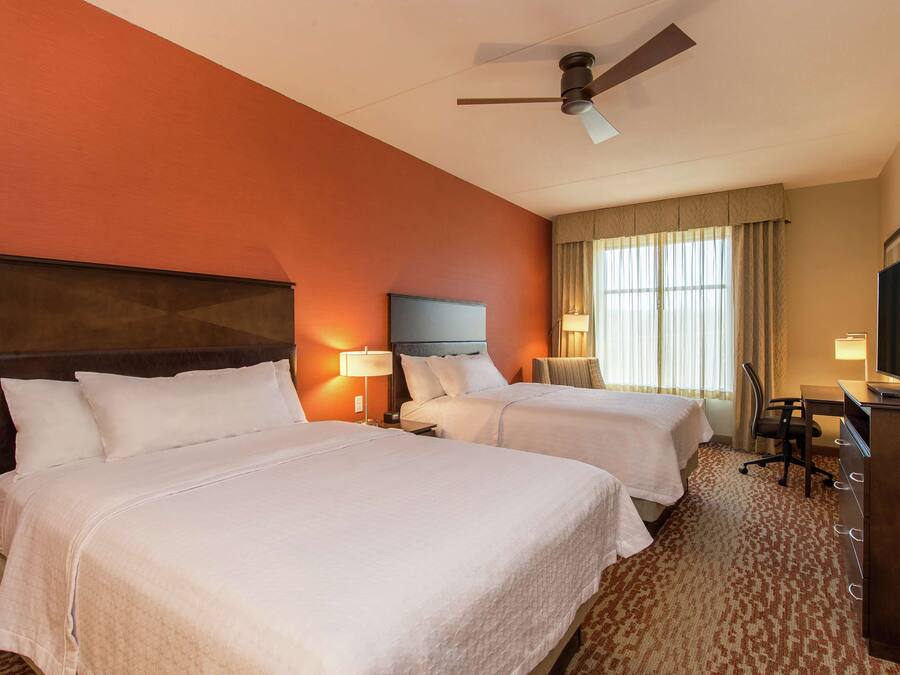Quarto de hotel com duas camas, uma escrivaninha, uma cadeira, uma televisão e um ventilador de teto.