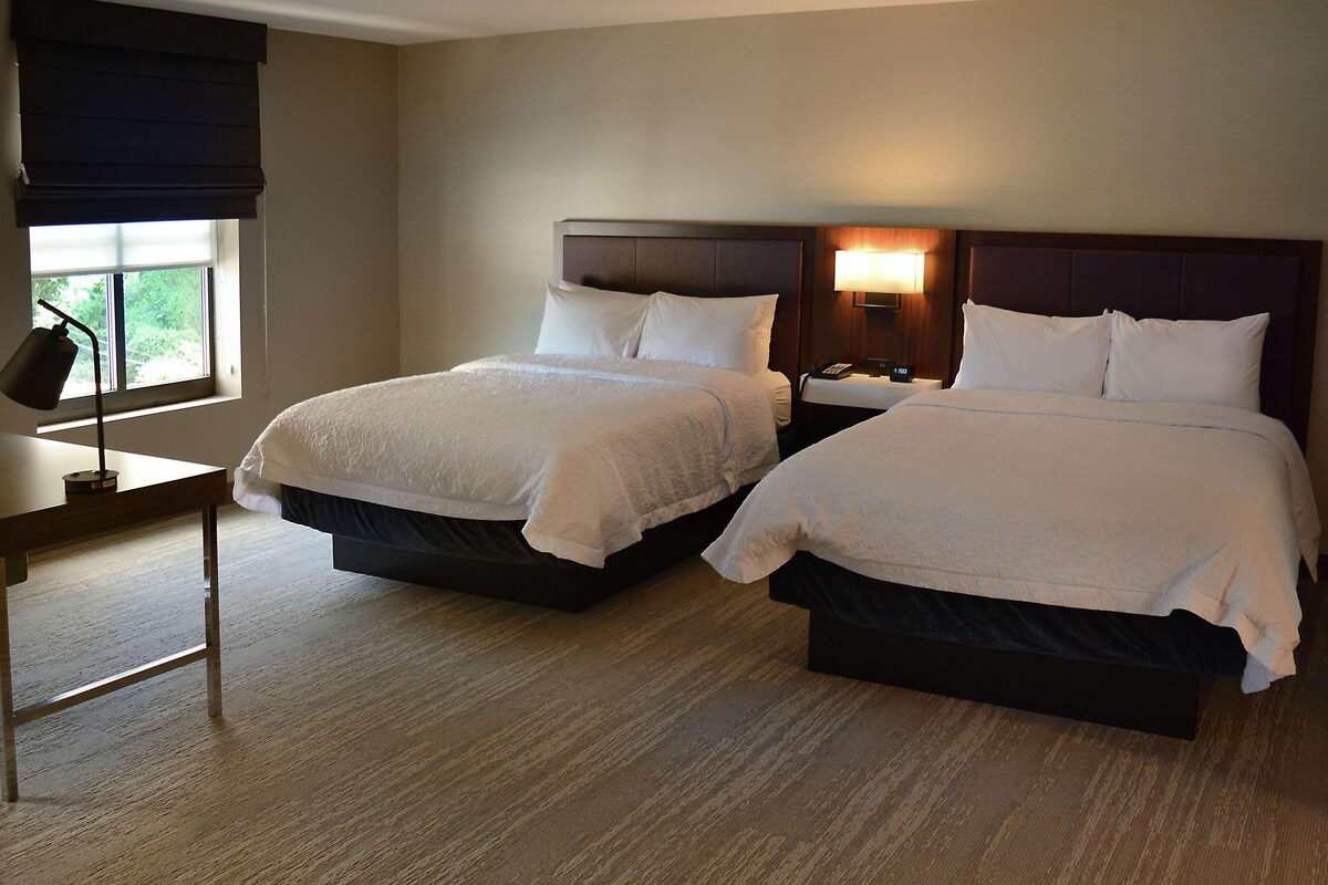 Quarto de hotel com duas camas, uma mesa de cabeceira com abajur e uma janela com vista.
