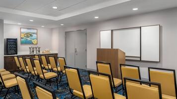 Uma sala de conferências com fileiras de cadeiras, um quadro branco, um espaço para café e um quadro na parede.