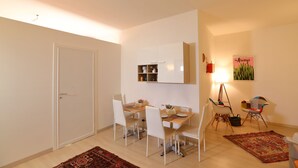 Uma sala de jantar com mesa posta para quatro, cadeiras brancas, tapete vermelho estampado e um aparador fixado na parede com objetos decorativos.