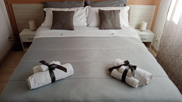 Uma cama bem arrumada com duas toalhas amarradas com fitas pretas, uma cabeceira cinza e uma mesa de cabeceira com abajur.
