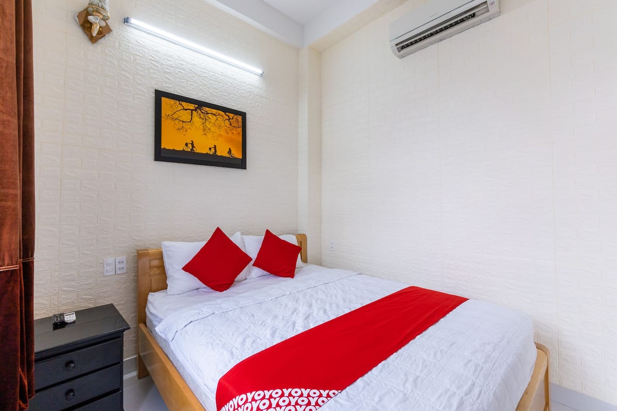Cama bem arrumada com colcha branca e vermelha, cabeceira de madeira e manta vermelha. Pequeno criado-mudo com abajur e tomada. Uma moldura com imagem pendurada na parede acima da cama.