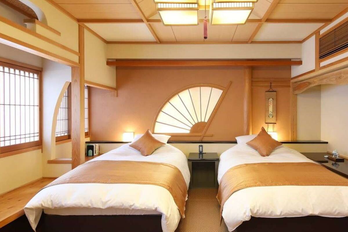 Quarto de hotel com duas camas, um ventilador, uma janela e teto no estilo tradicional japonês.