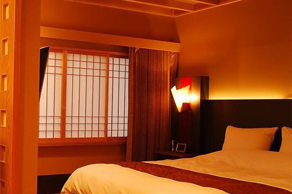 Quarto de hotel com uma cama grande, uma mesa de cabeceira, uma luminária e uma janela com persianas.