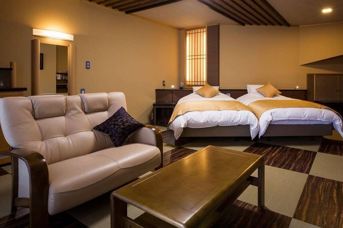 Quarto de hotel com sofá, duas camas, mesa de cabeceira, abajur e mesa de centro de madeira.