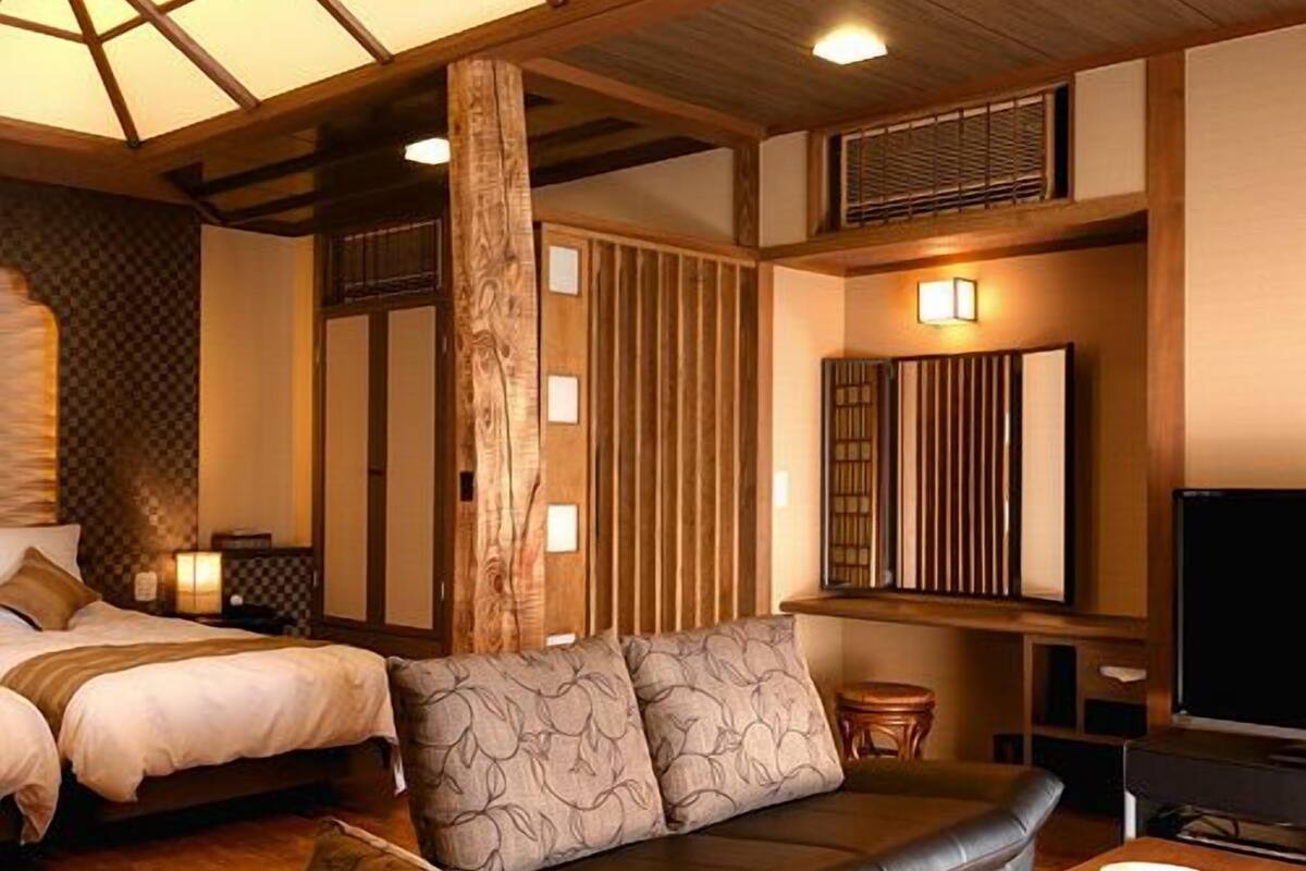 Quarto de hotel com cama, sofá, televisão e teto de madeira.