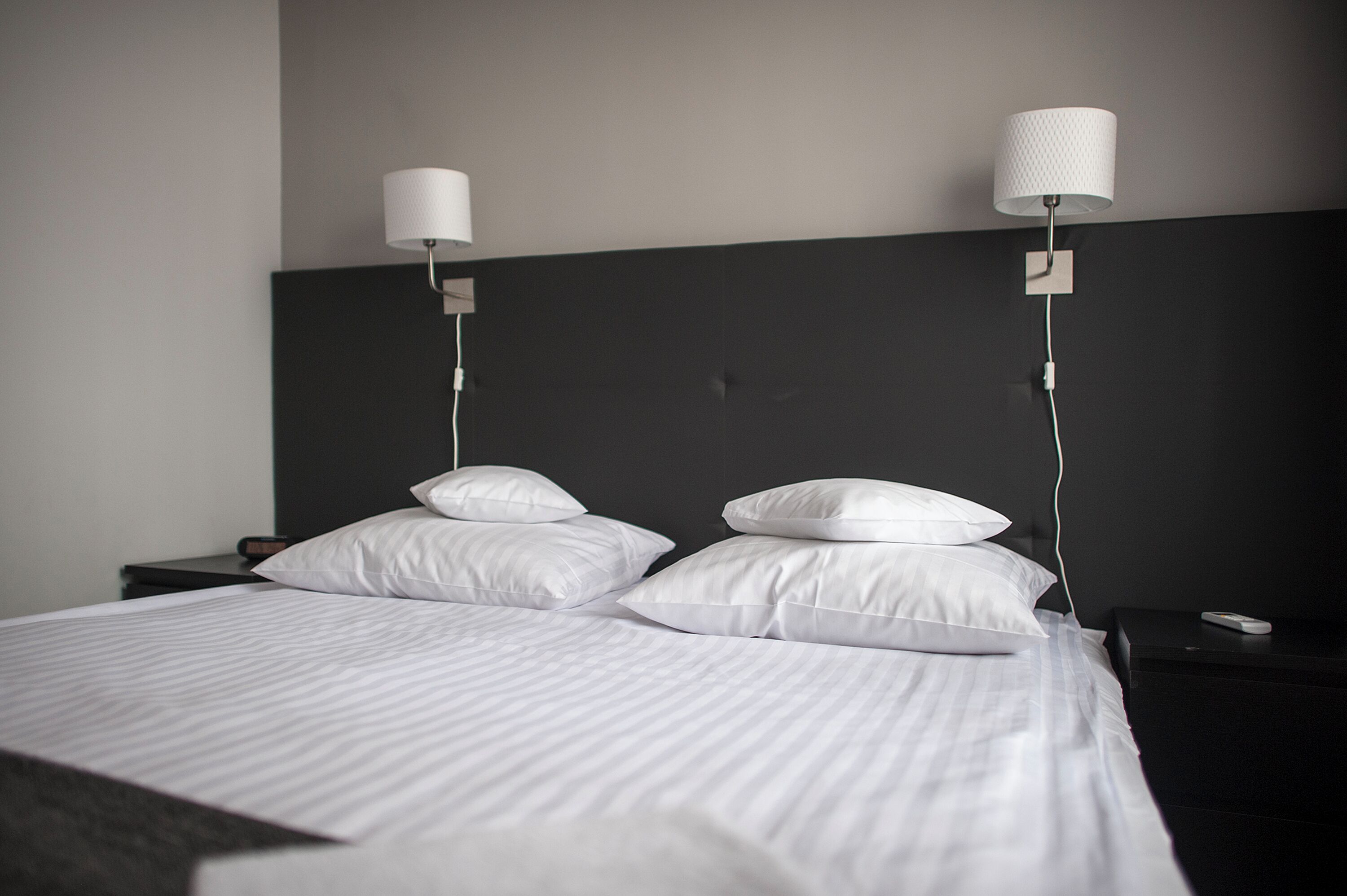 Cama bem arrumada com lençóis e almofadas brancas, duas mesas de cabeceira com abajures e uma cabeceira escura.