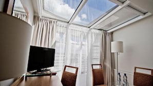 Quarto de hotel com televisão, duas cadeiras, uma mesa, abajur e vista do céu por uma grande janela.