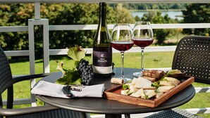 Uma mesa com uma garrafa de vinho, duas taças de vinho tinto, uma tábua de queijos e uma garrafa de champanhe em uma varanda com vista para um lago.